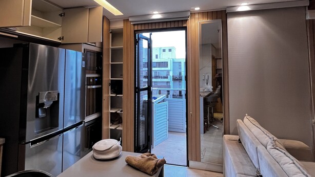 Uma cozinha moderna com frigorífico embutido, área de refeições com mesa e cadeiras, e vista para um prédio residencial através de uma porta aberta.