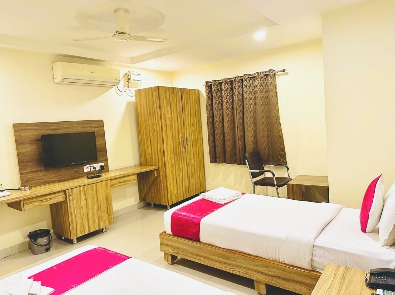 Quarto de hotel com escrivaninha de madeira, televisão, cama com roupa de cama vermelha e branca, cadeira e ar condicionado.