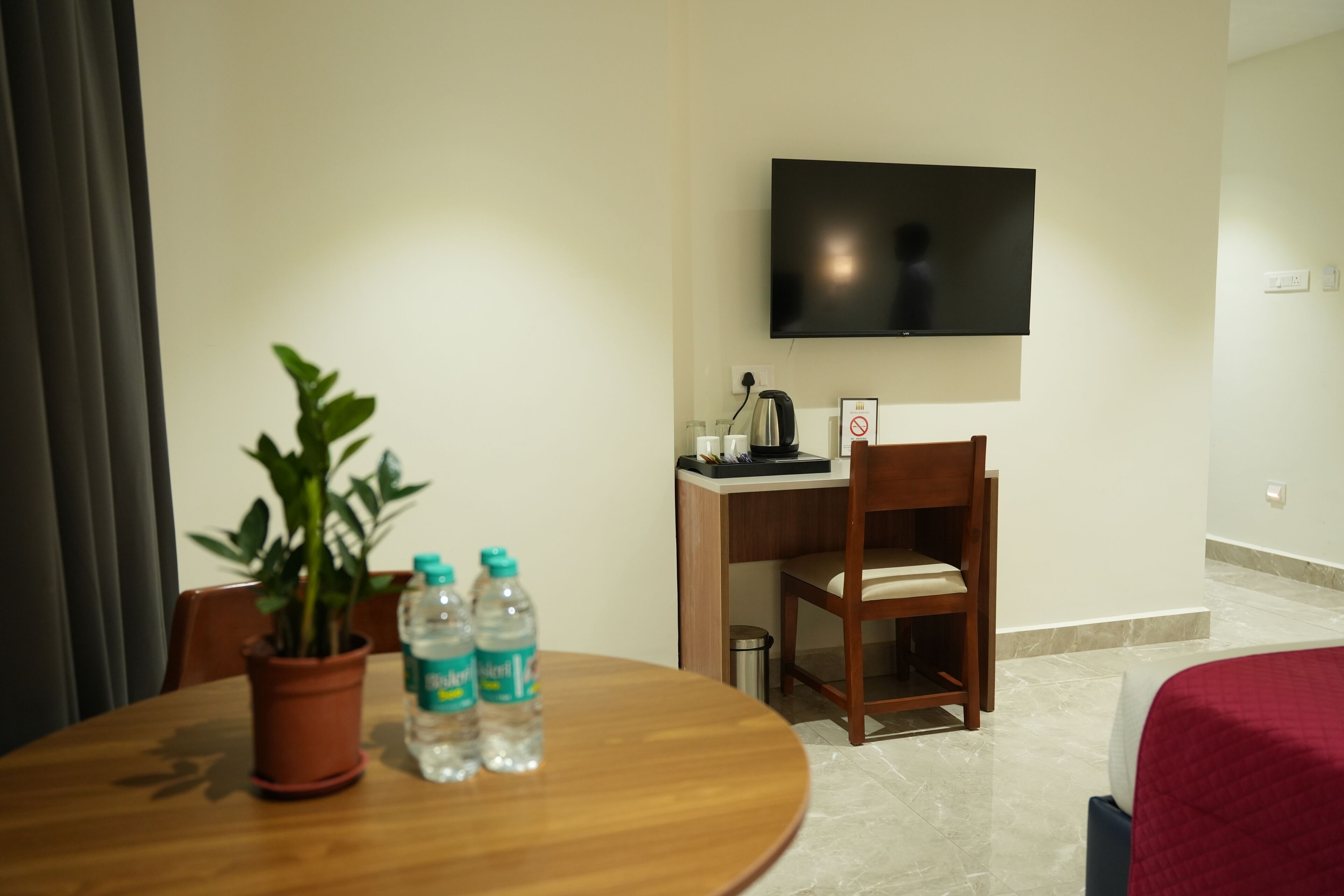 Quarto de hotel com uma mesa de jantar de madeira, duas garrafas de água, um vaso de plantas, uma TV fixada na parede, uma escrivaninha com cadeira e uma cama com um cobertor vermelho.