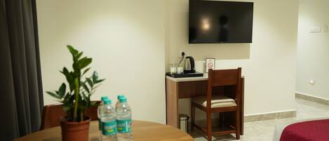 Quarto de hotel com uma mesa de jantar de madeira, duas garrafas de água, um vaso de plantas, uma TV fixada na parede, uma escrivaninha com cadeira e uma cama com um cobertor vermelho.