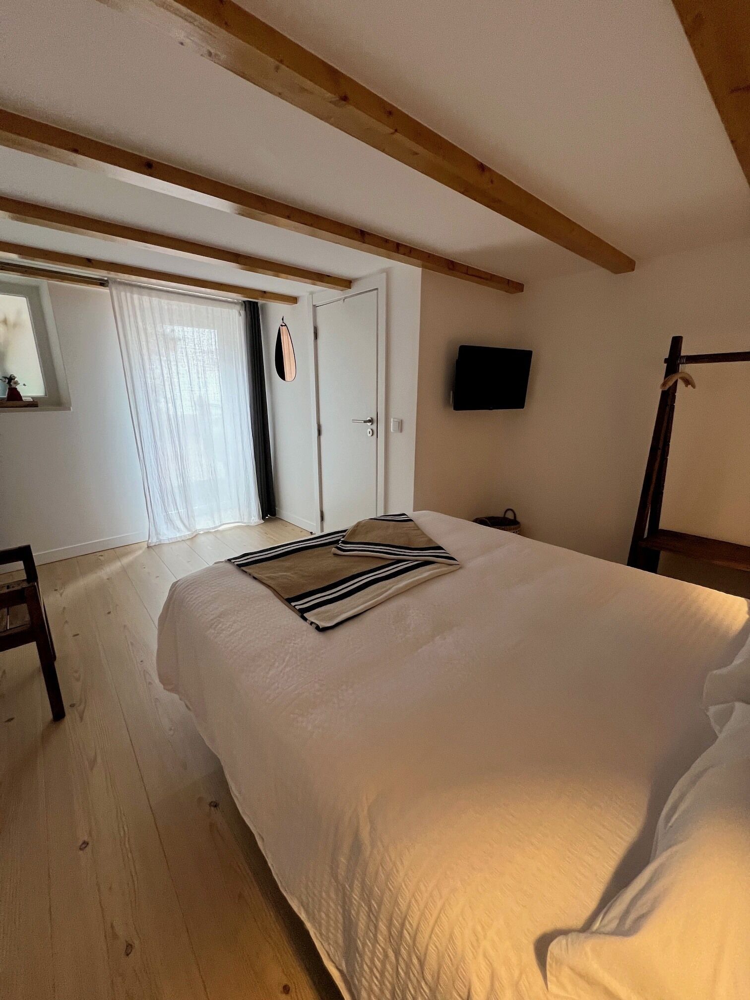 Quarto com cama grande, piso de madeira, porta, janela com cortinas e TV fixada na parede.