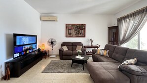 Uma sala de estar com televisão de tela plana, um ventilador de chão, um sofá, uma mesa de centro e um quadro na parede.