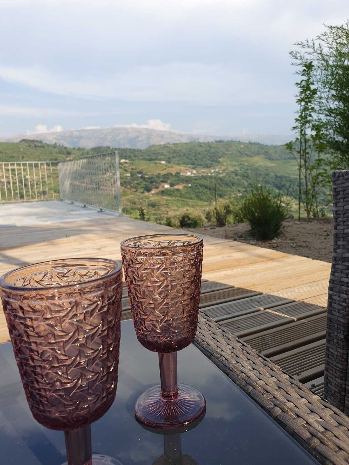 Dois copos de vidro sobre uma mesa, com vista para uma paisagem montanhosa.