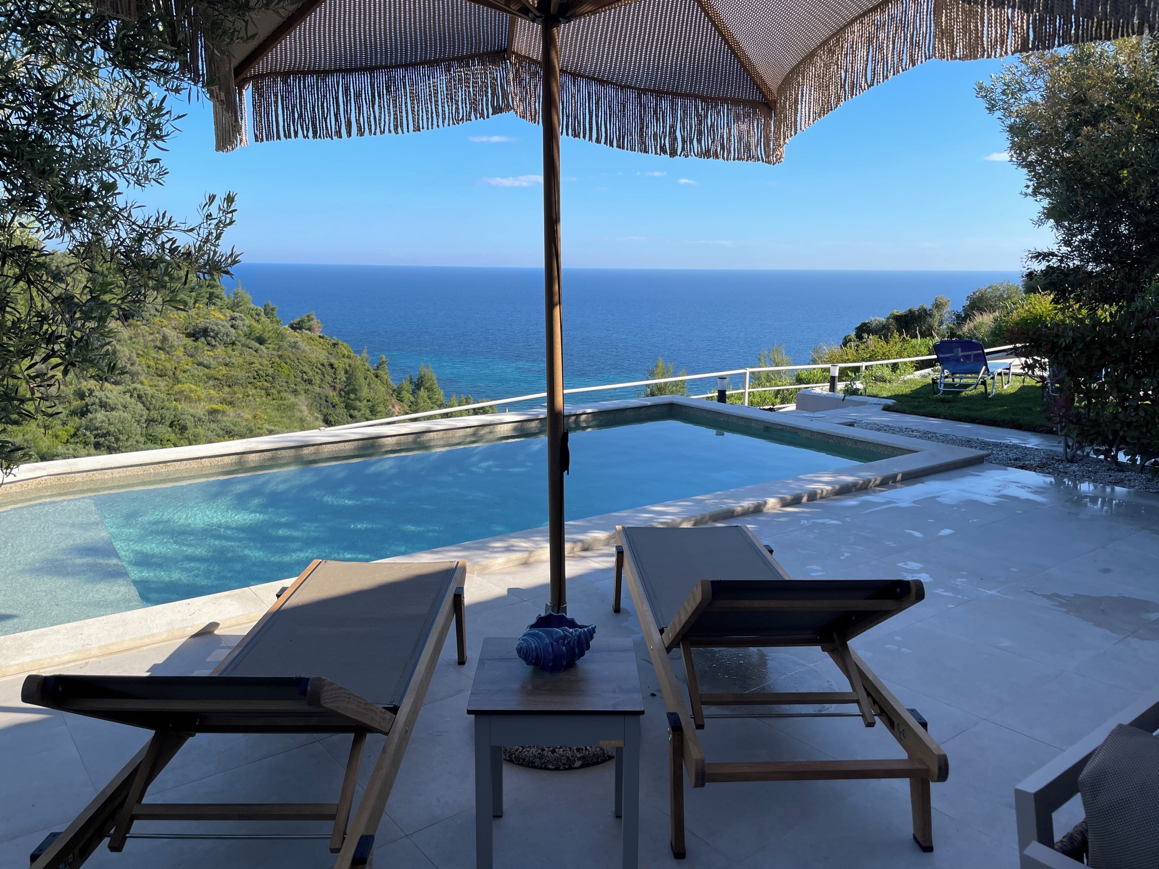 Uma área à beira da piscina com duas espreguiçadeiras, uma mesa e vista para o mar.