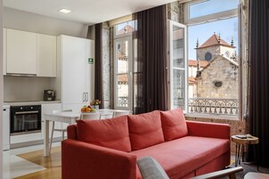 Uma sala de estar moderna com um sofá vermelho, uma mesa de jantar com cadeiras e uma cozinha com armários e eletrodomésticos brancos. É visível uma varanda com grade de ferro e vista para um edifício histórico pela janela.