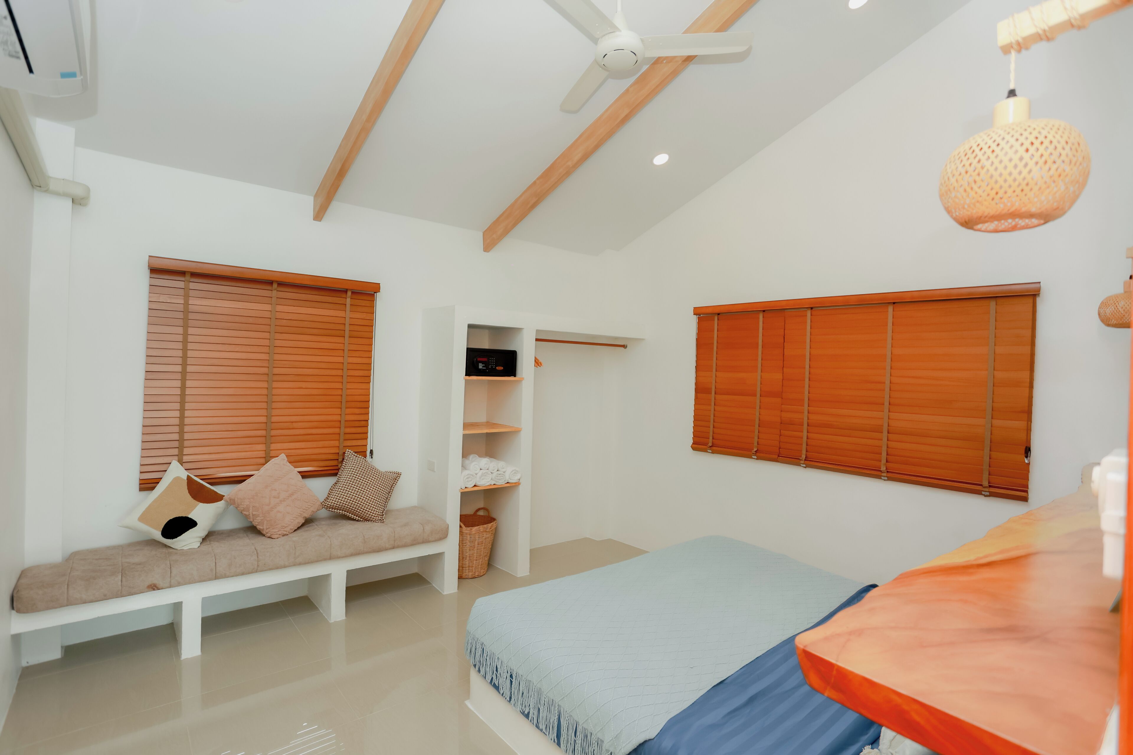 Quarto moderno com cama, um banquinho com almofadas, persianas de madeira e ventilador de teto.