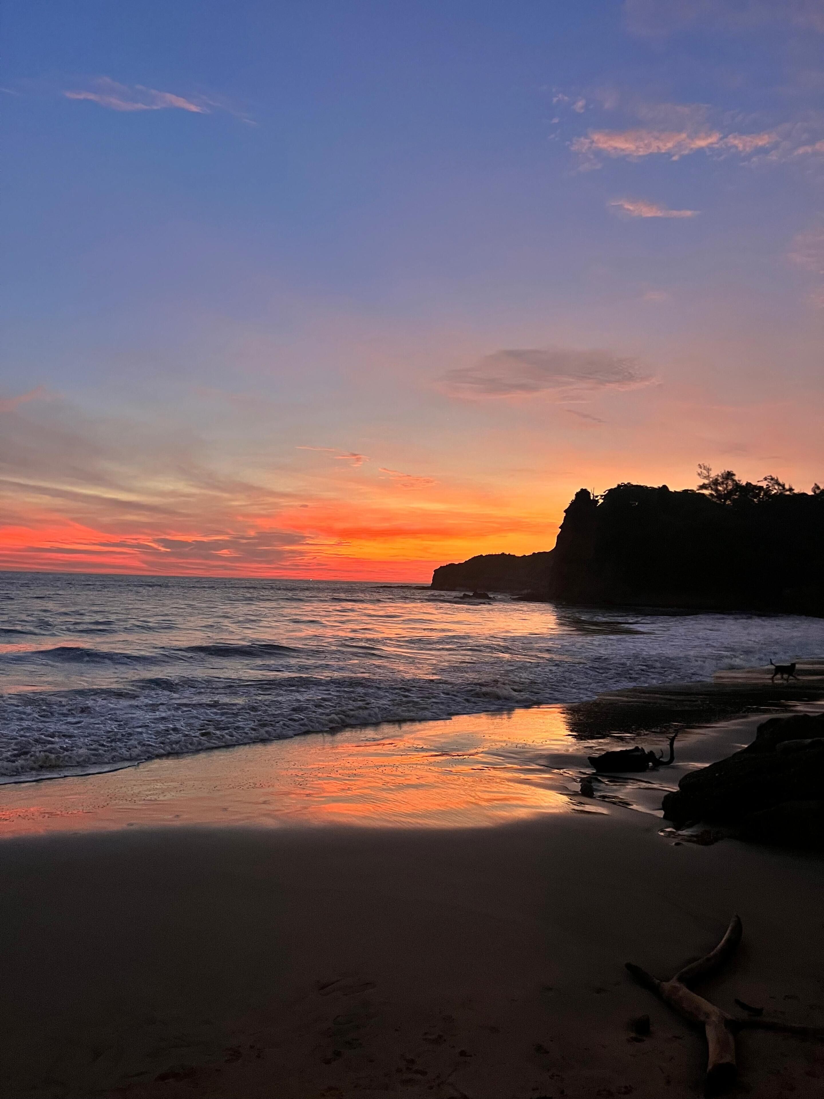 Uma praia com ondas, um pôr do sol e um penhasco.