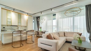 Uma sala de estar moderna com um sofá, mesa de centro e banquetas. Há uma área de cozinha com bancada de mármore e armários.