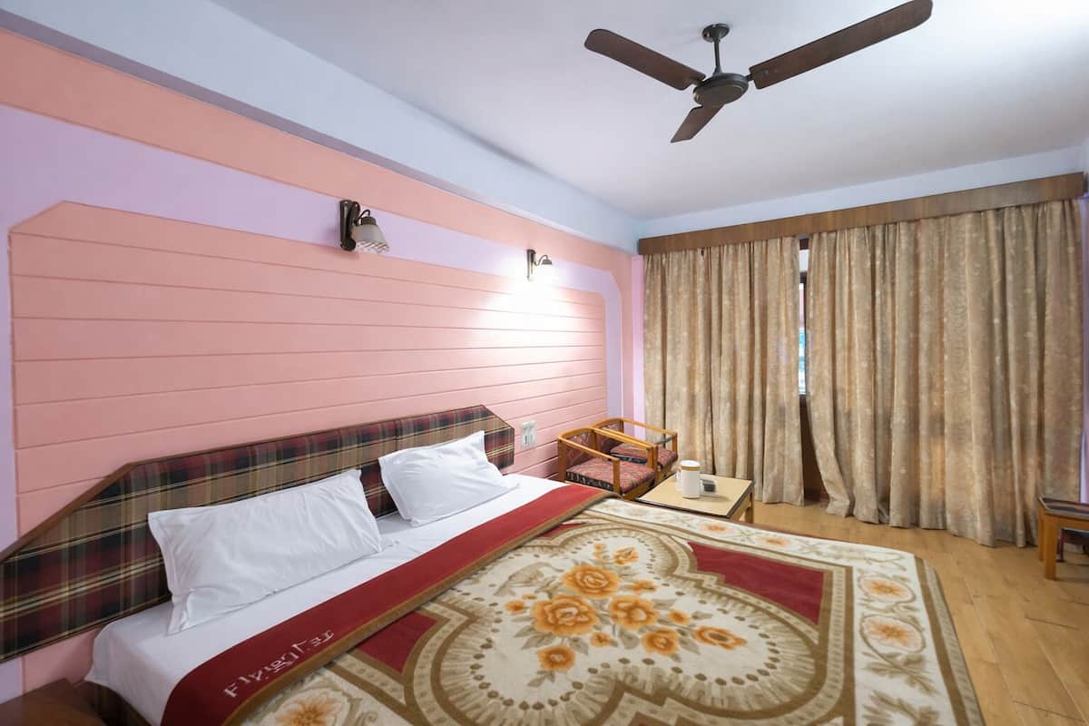 Um quarto com cama, ventilador de teto, janela com cortinas e uma pequena mesa.