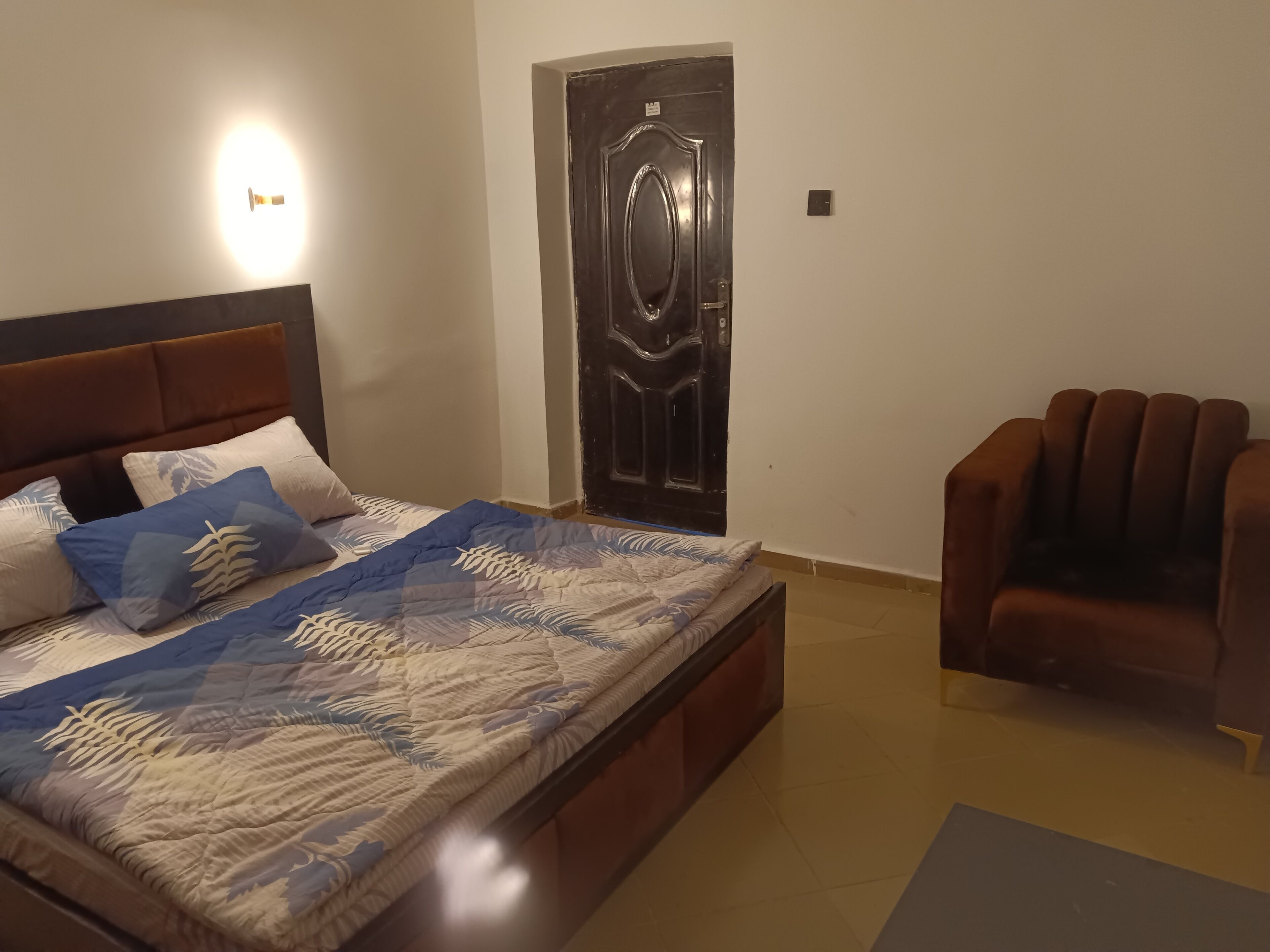 Um quarto com uma cama, uma cadeira, uma porta e uma luminária fixada na parede.