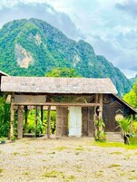 Um edifício rústico de madeira com telhado de palha, rodeado por vegetação exuberante e montanhas ao fundo.