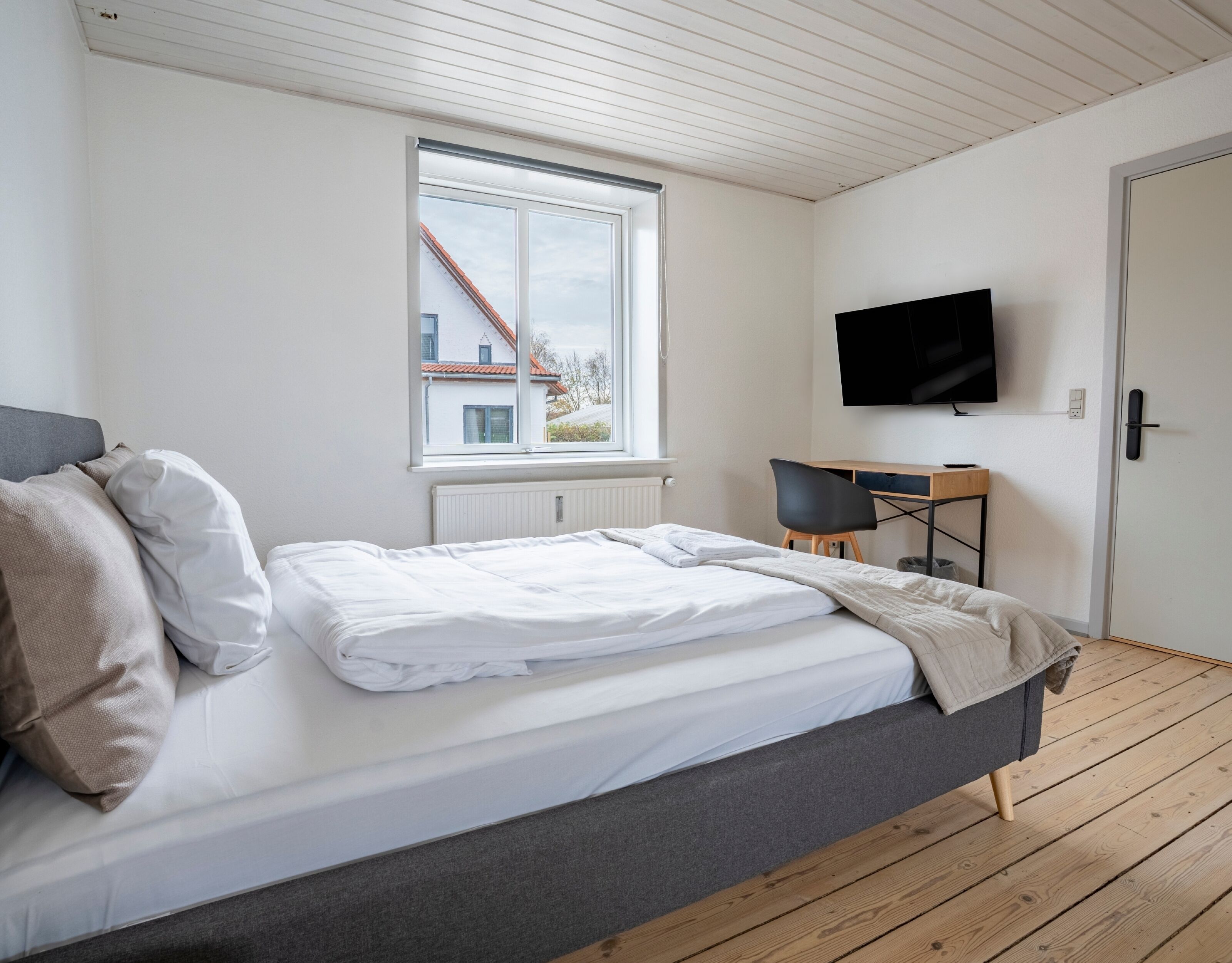 Quarto com cama, escrivaninha, cadeira, televisão e janela com vista para uma casa.