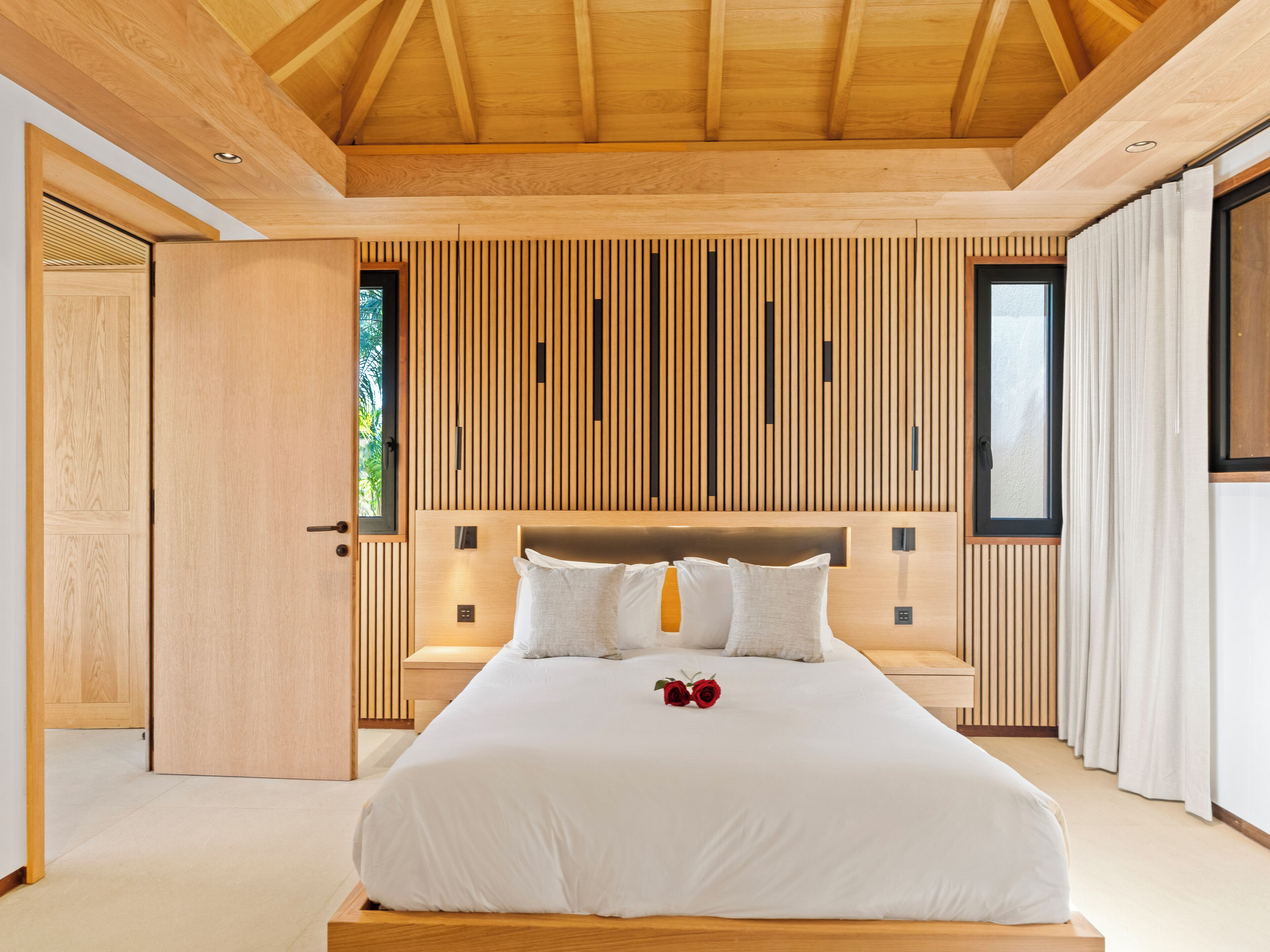 Quarto moderno com teto de madeira, uma cama grande com roupa de cama branca e uma janela com cortinas.