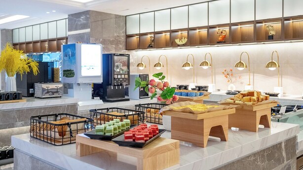 Um hall de hotel moderno, com uma recepção de mármore, um painel digital e diversas opções de comida e bebida.