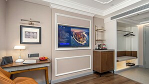 Quarto de hotel com piso de madeira, uma escrivaninha com luminária, um quadro emoldurado, uma tela digital, um guarda-roupa e um espaço de estar com um banco.