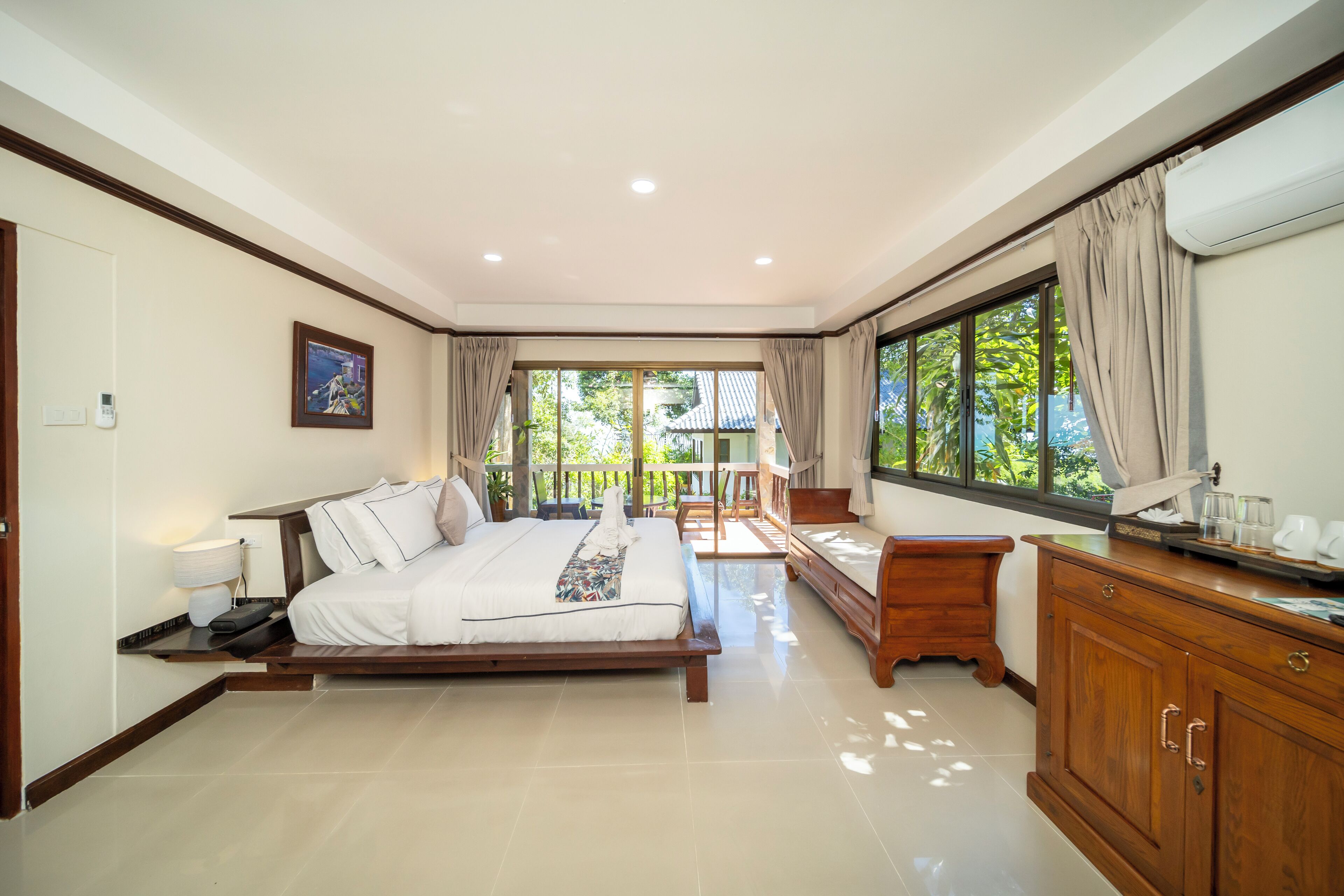 Um quarto espaçoso com uma cama grande, uma mesa de cabeceira de madeira, um aparador de madeira e vista para o exterior por grandes janelas.