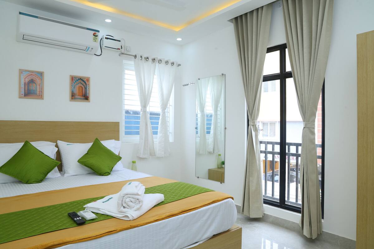 Quarto de hotel com cama, almofadas e colcha verdes, espelho, janela com cortinas e vista para o exterior.