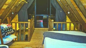 Quarto no sótão com teto inclinado, vigas de madeira, luminária central e cama com edredom branco.