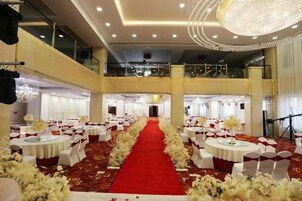 Uma sala de banquetes com mesas redondas, tapete vermelho e arranjos florais brancos.