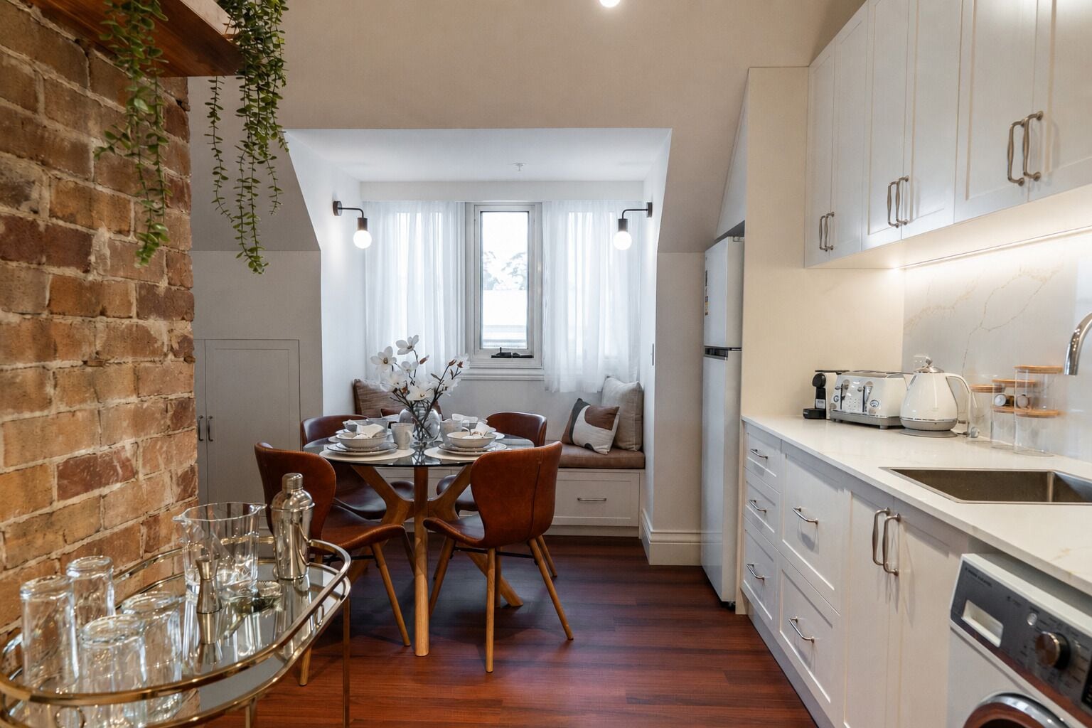 Uma cozinha compacta com armários brancos, uma mesa de jantar com cadeiras e uma janela com cortinas.