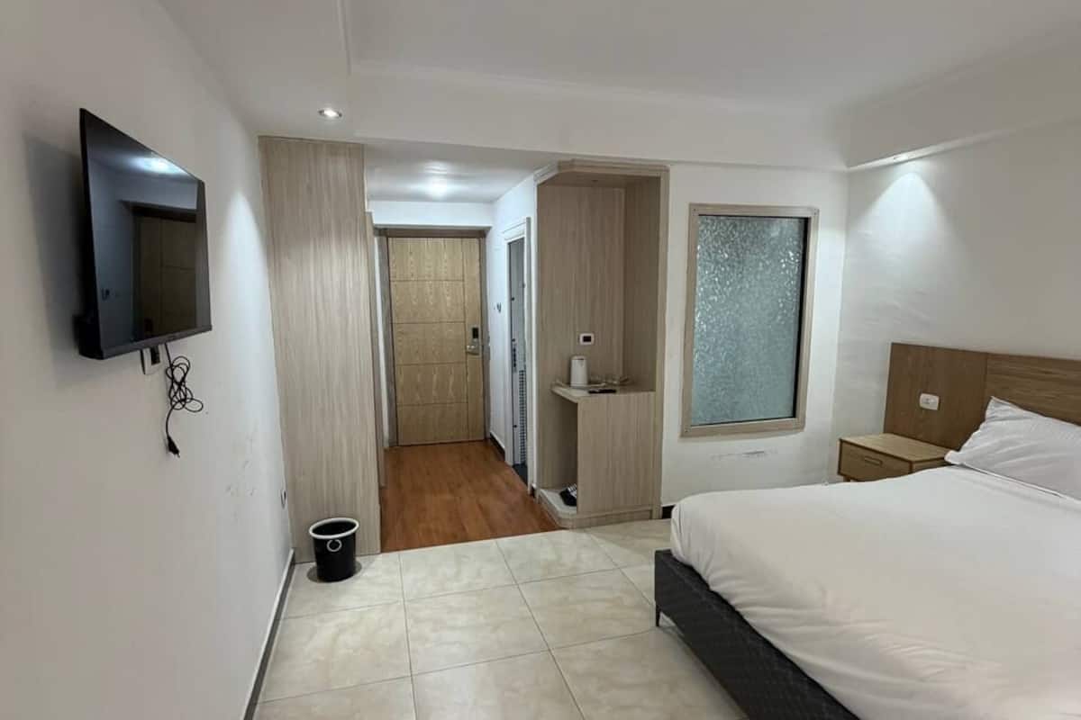 Quarto de hotel com uma cama, uma TV fixada na parede, uma janela e uma porta que dá para um corredor.