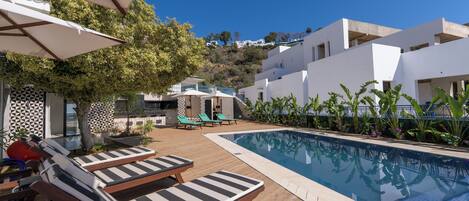 Área junto à piscina com espreguiçadeiras, piscina e vista para uma encosta com edifícios.