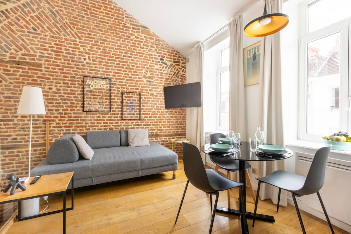 Uma sala de estar moderna com parede de tijolos, um sofá cinza, uma mesa de jantar com cadeiras e uma TV fixada na parede.