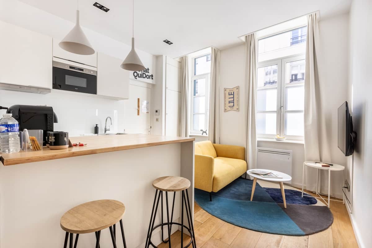 Cozinha moderna com micro-ondas, forno e máquina de café. Na sala de estar, há um sofá amarelo e uma pequena mesa redonda.
