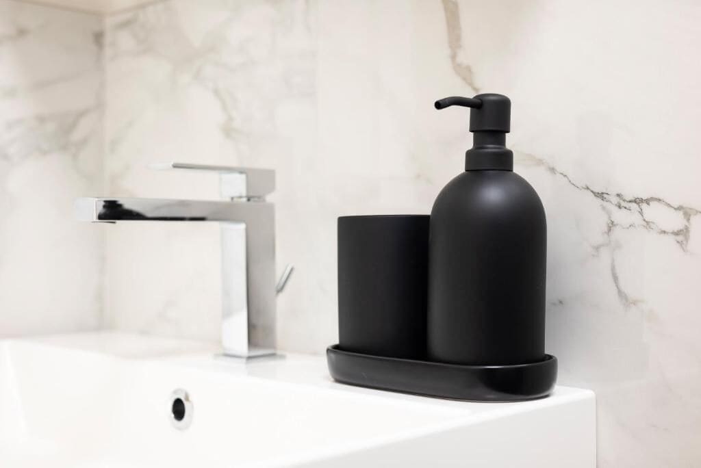 Casa de banho com lavatório branco, dispensador de sabonete preto e parede de mármore.