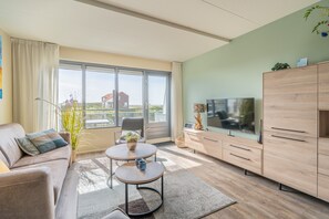 Uma sala de estar moderna com um sofá, duas mesas redondas, uma televisão e uma janela grande com vista para uma casa.