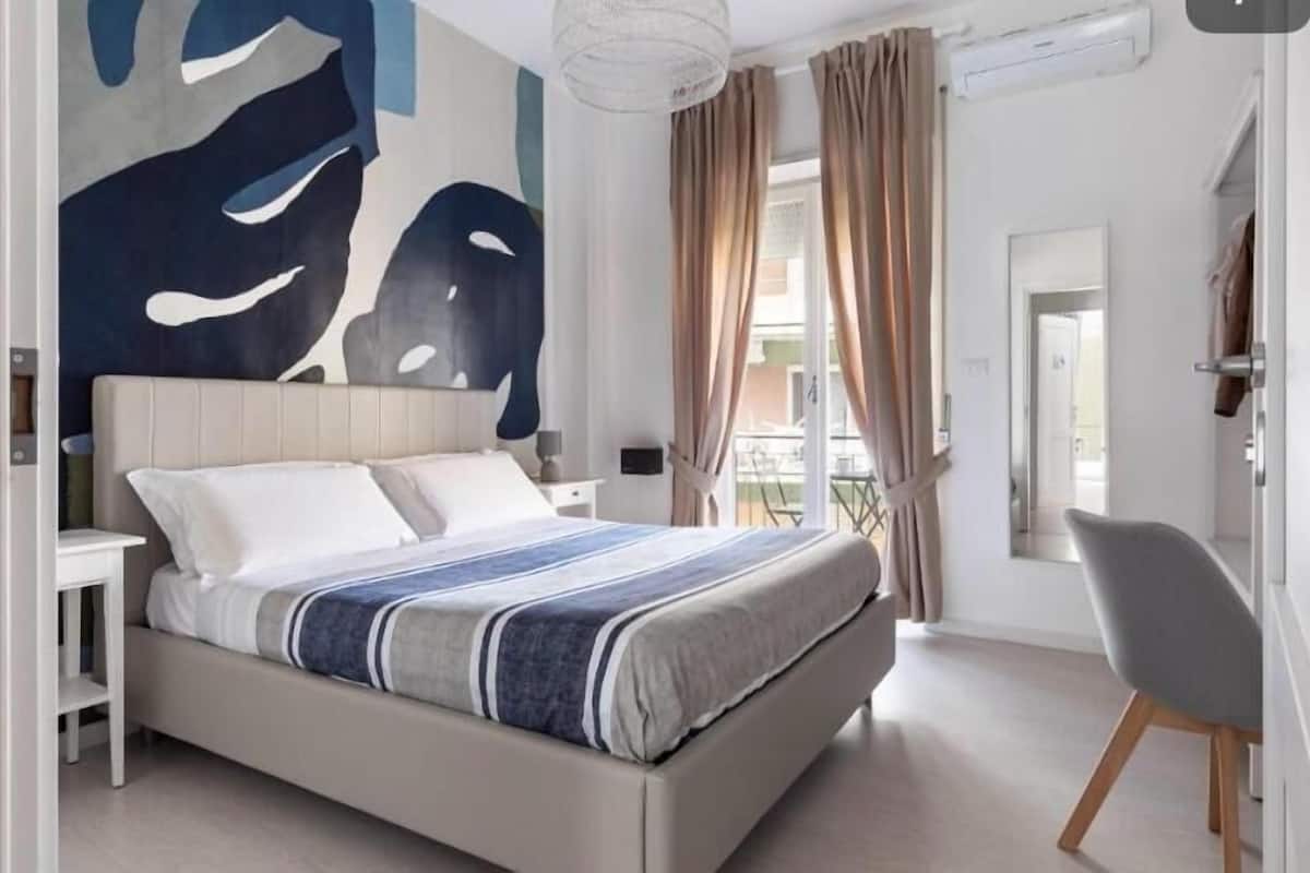 Quarto moderno com uma cama grande, uma mesinha de cabeceira, uma cadeira e vista para o exterior.