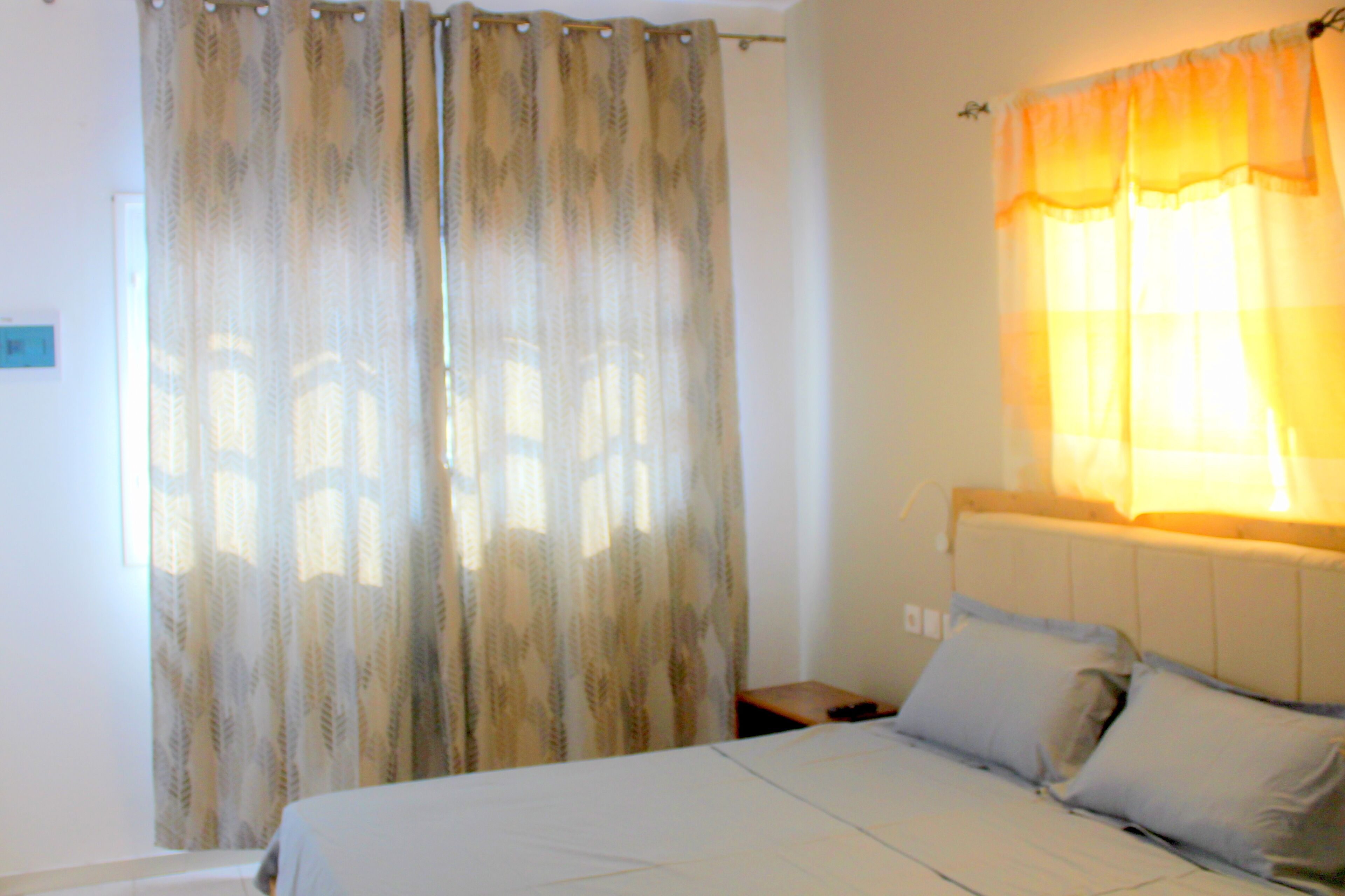 Um quarto com cama, almofadas, mesa de cabeceira e janela com cortinas.