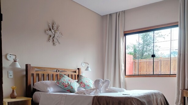 Um quarto com cama de madeira, roupa de cama branca, um objeto decorativo na parede e uma janela com cortinas.