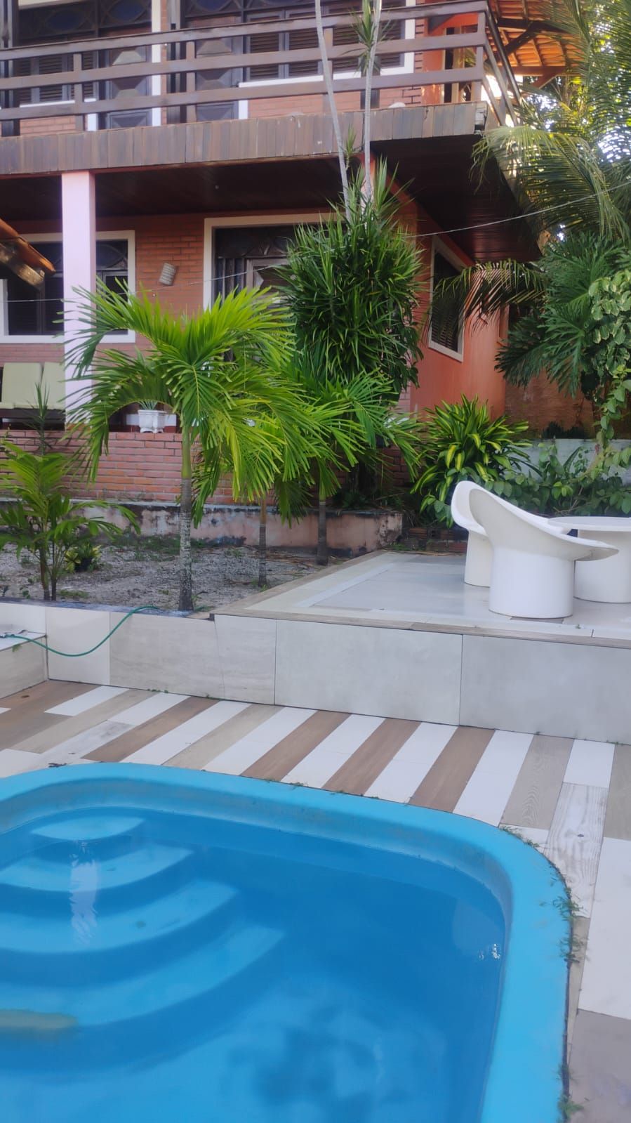 Piscina com deck de madeira, rodeada por plantas em vasos e um edifício com varanda.