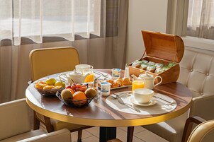 Mesa de café da manhã com diversos alimentos, incluindo frutas, doces e bebidas, em um ambiente bem iluminado com vista para o exterior.