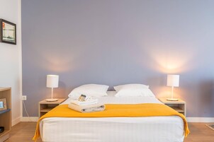 Uma cama bem arrumada com lençóis brancos e um cobertor amarelo, ladeada por duas mesas de cabeceira com luminárias, em um quarto com parede azul claro.