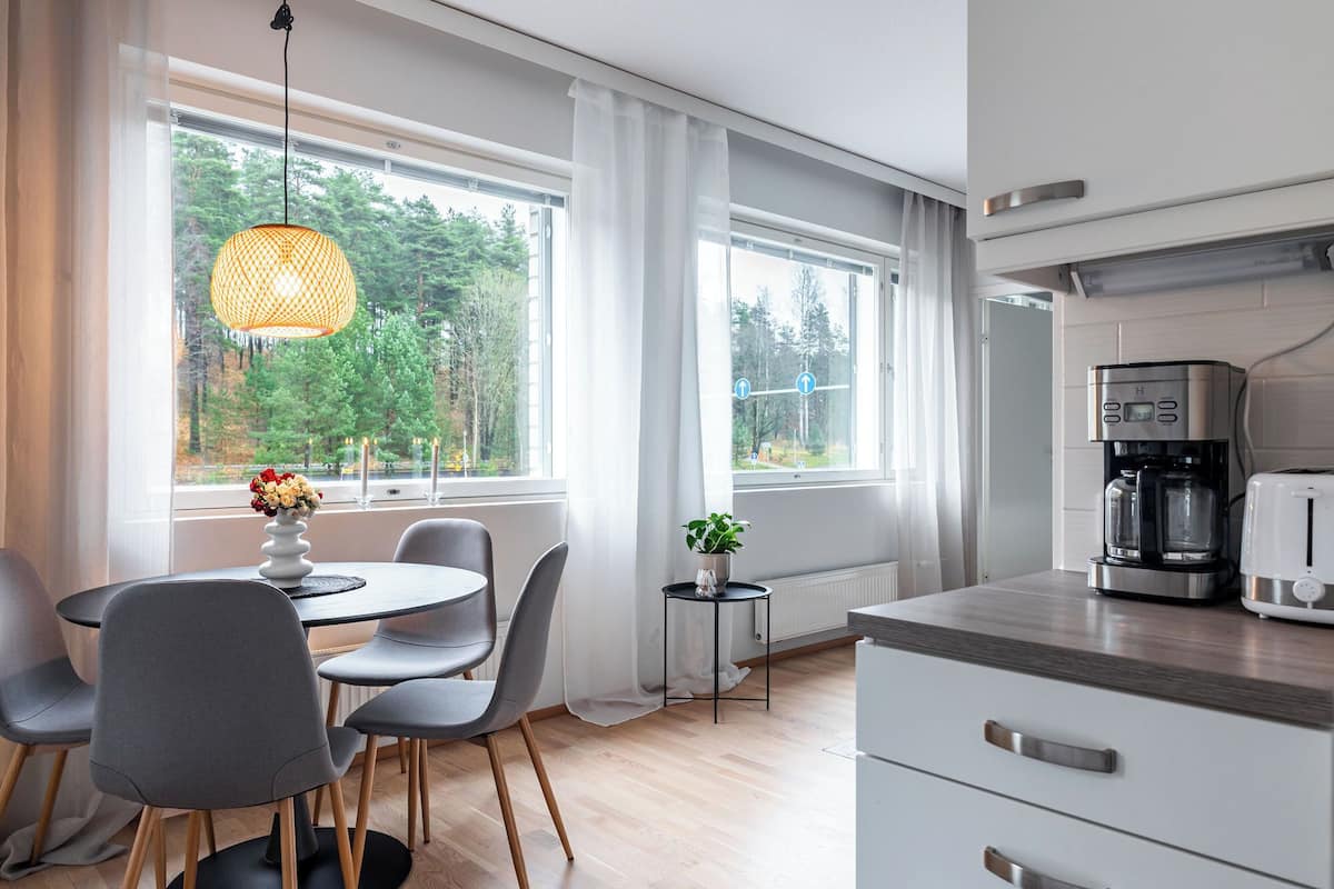 Cozinha moderna com cafeteira, área de refeições com mesa e cadeiras, e janelas amplas com vista para árvores.