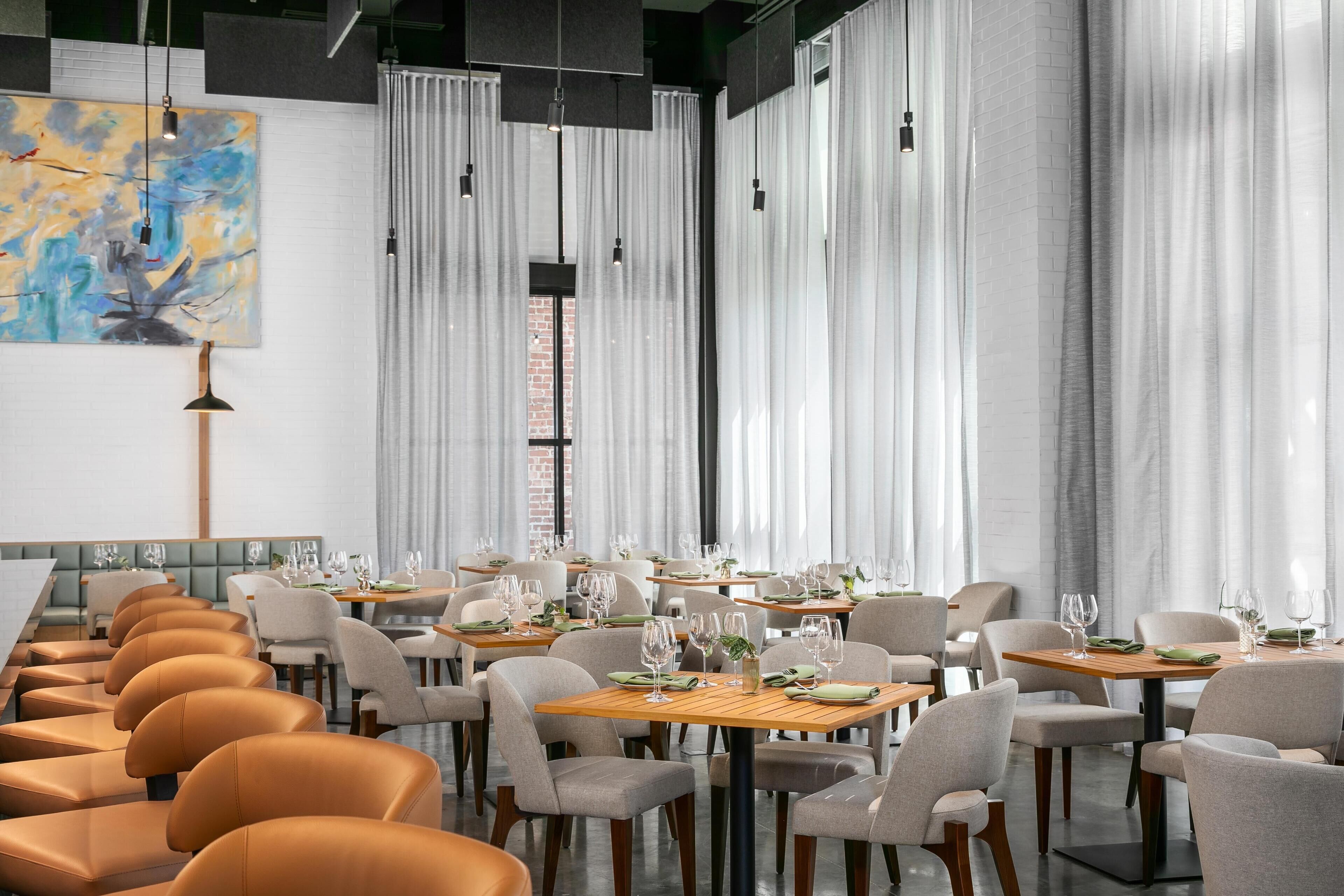 Um restaurante com várias mesas e cadeiras, uma parede de tijolos e uma grande pintura abstrata.