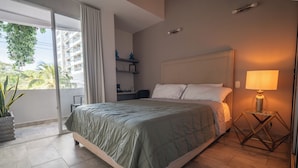 Quarto de hotel com cama, mesa de cabeceira com luminária, planta em vaso, porta de correr para uma varanda e vista de edifícios e vegetação lá fora.