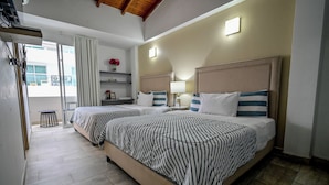 Quarto de hotel com duas camas, uma varanda, uma mesinha e uma prateleira na parede com flores.