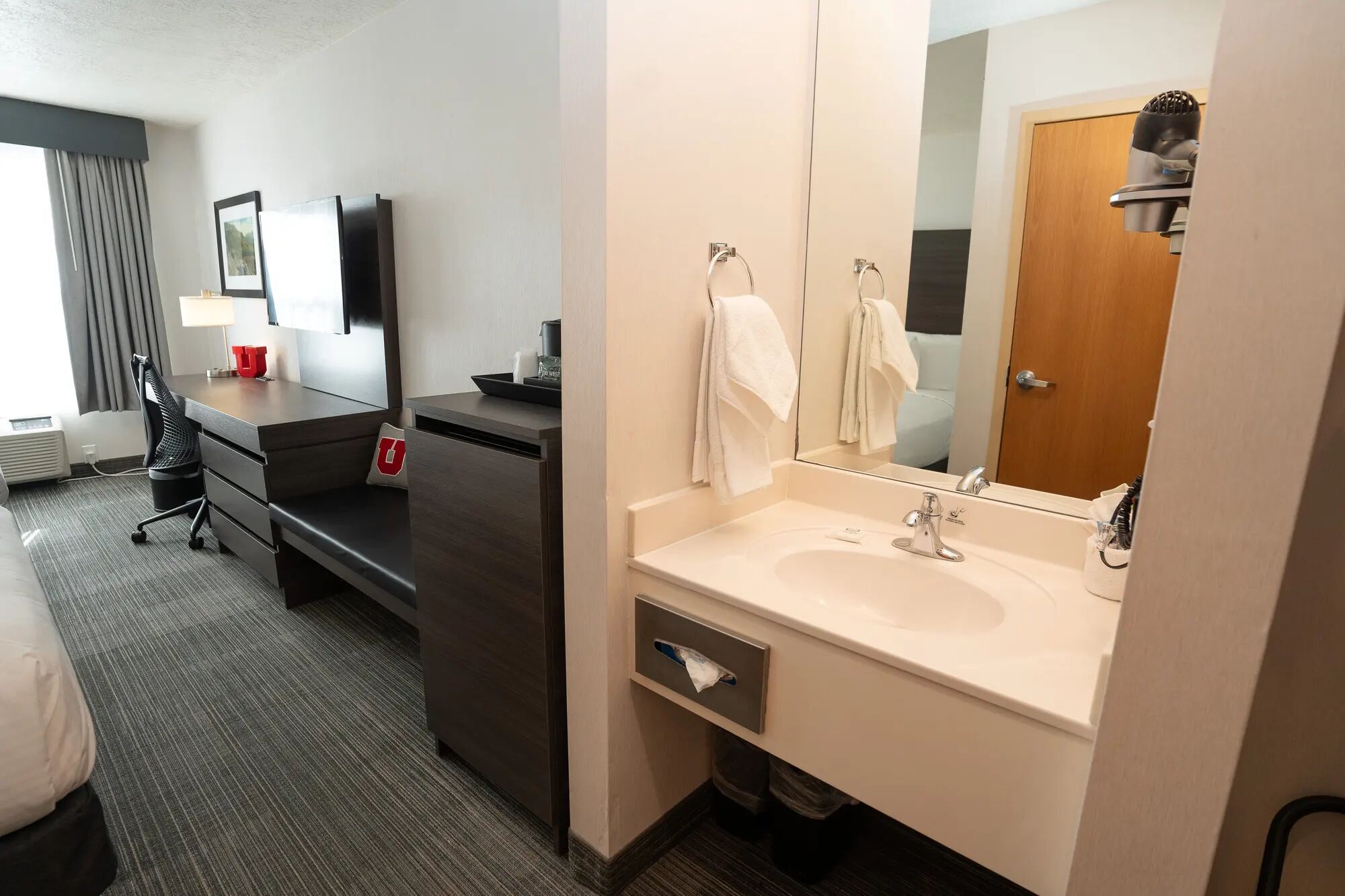 Quarto de hotel com uma cama, uma mesa com cadeira, luminária de mesa, televisão de tela plana, espelho grande, pia branca e porta de madeira.
