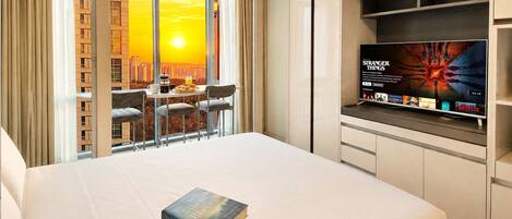 Quarto de hotel com cama, televisão exibindo um filme, varanda com vista para a cidade e mesa com itens de café da manhã.