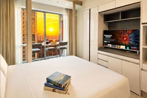 Quarto de hotel com cama, televisão exibindo um filme, varanda com vista para a cidade e mesa com itens de café da manhã.
