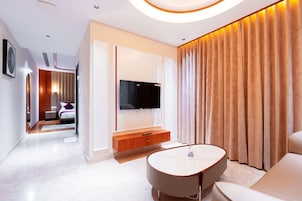 Quarto de hotel moderno com televisão de tela plana, mesa de centro redonda, sofá e uma grande janela com cortinas.