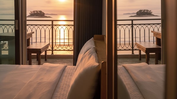 Quarto de hotel com uma cama, duas cadeiras de madeira e vista para o pôr do sol sobre a água.