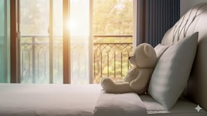 Uma cama com um ursinho de pelúcia, uma janela com vista para uma varanda e um quarto iluminado pelo sol.