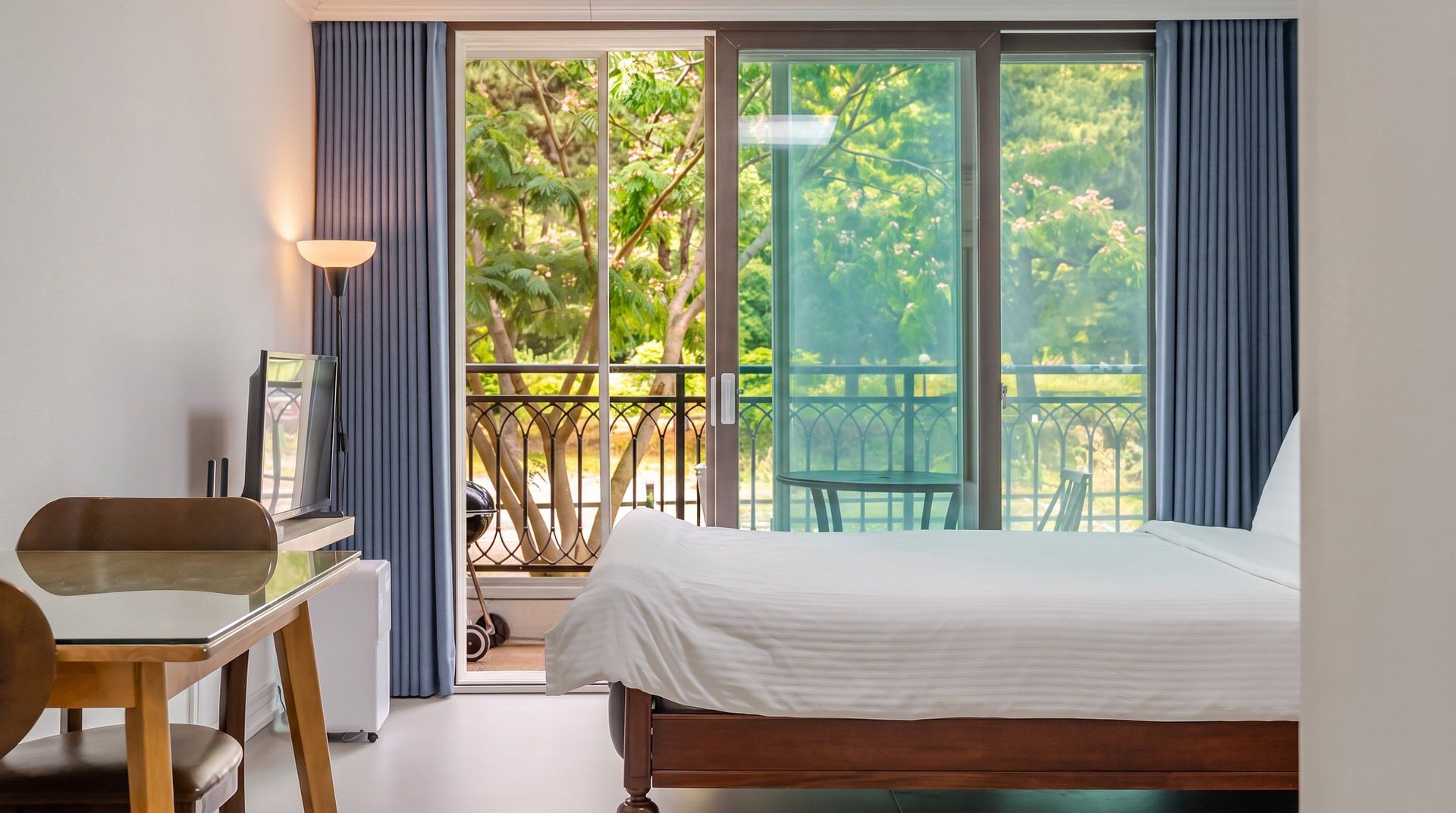 Quarto com cama, escrivaninha e cadeira. Possui varanda com vista para árvores e corrimão.