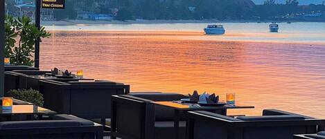Restaurante com mesas ao ar livre, vista para a água, uma placa e colinas ao longe.