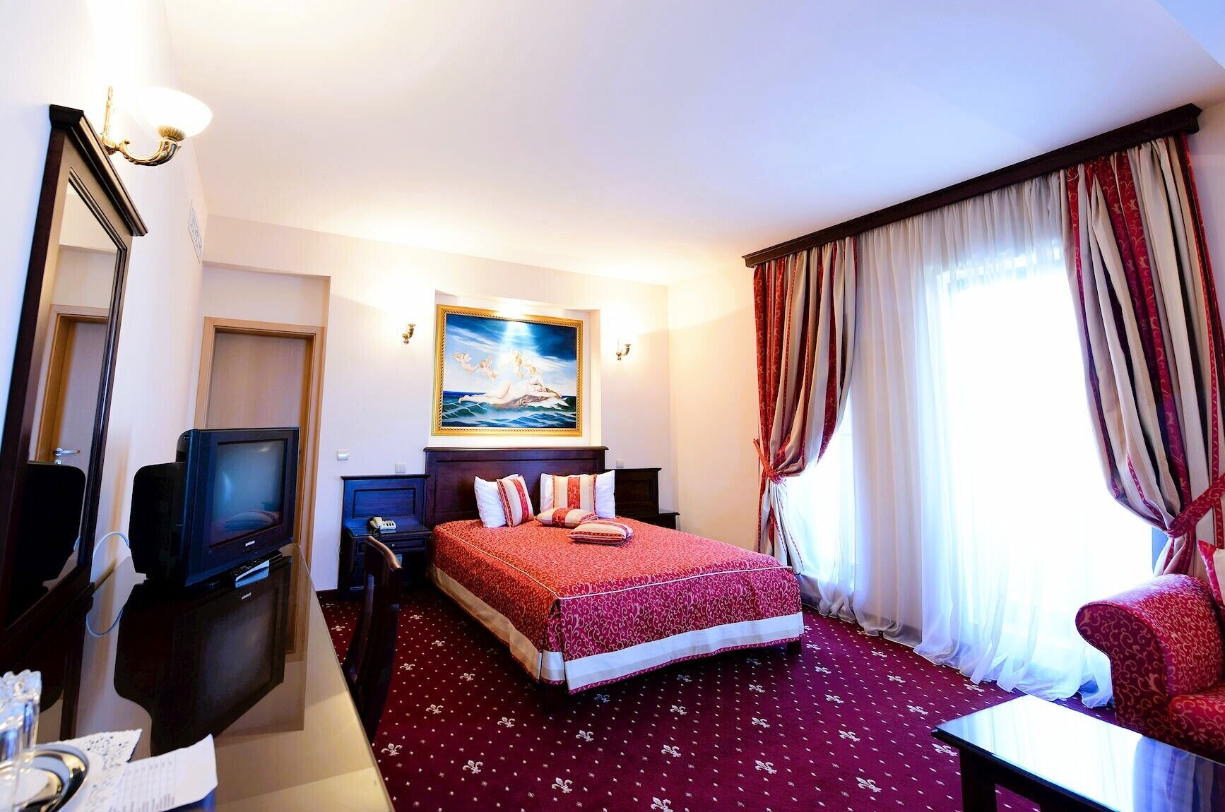 Quarto de hotel com cama, televisão, escrivaninha e um quadro na parede.