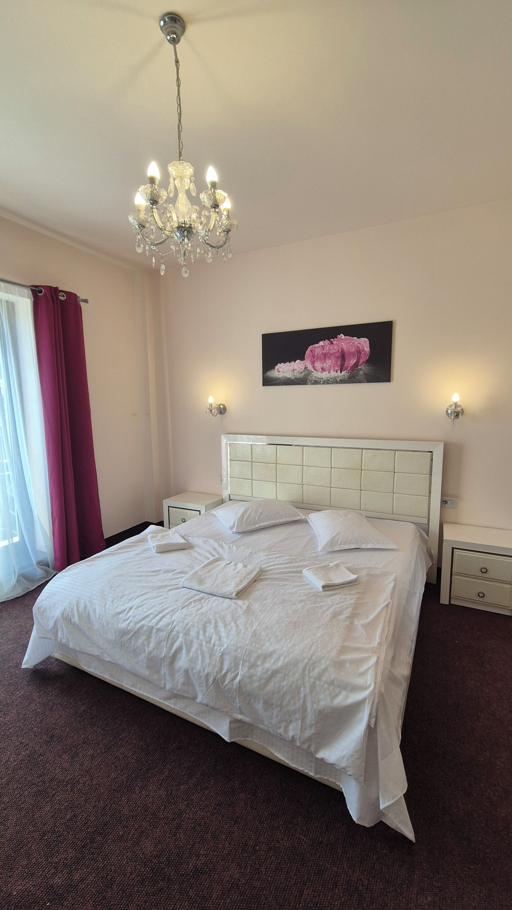 Uma cama bem arrumada com roupa de cama branca, um candelabro, um quadro com flores cor-de-rosa e uma cortina vermelha.
